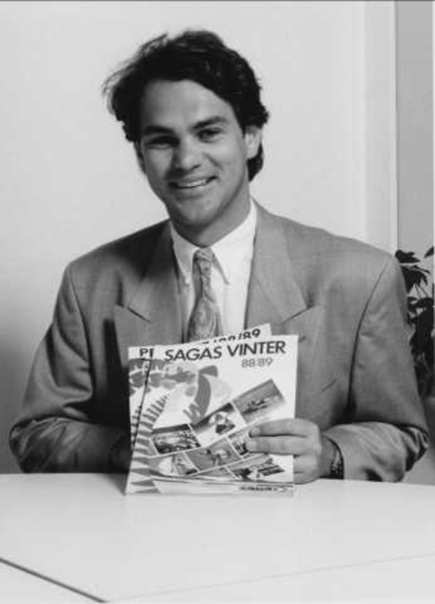 Per G. Braathen sittende bak bord. Holder fram brosjyrer for Saga ...