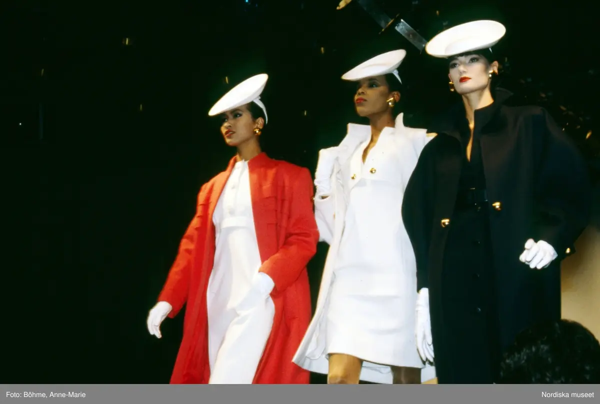 Modevisning. Modeller i klänningar och kappor i rött, vitt och svart ...