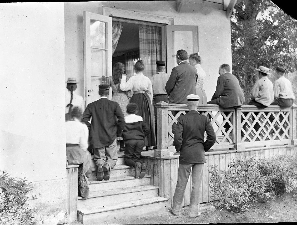 Ett sällskap med vuxna och unga står och sitter på en veranda till ett ...
