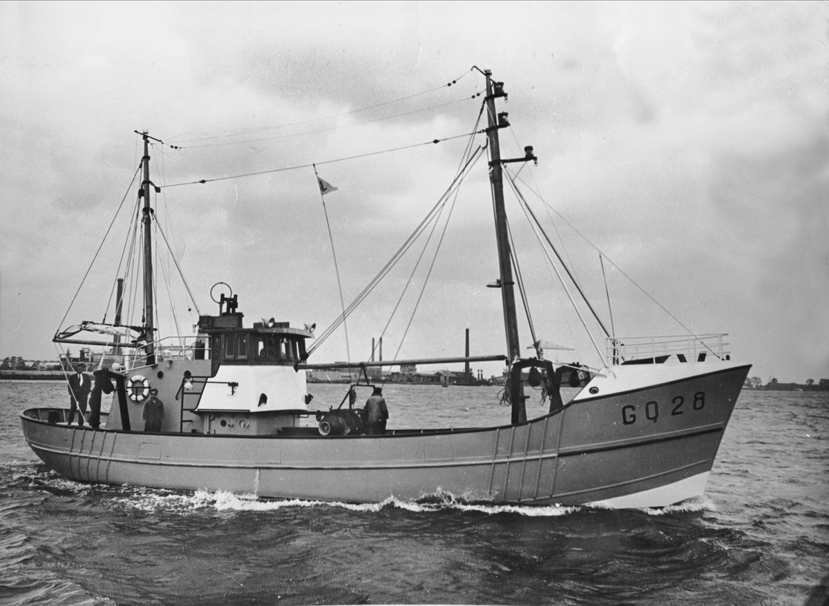 Fiskekutter i fart, med mannskap på dekk og bebyggelse i bakgrunnen ...