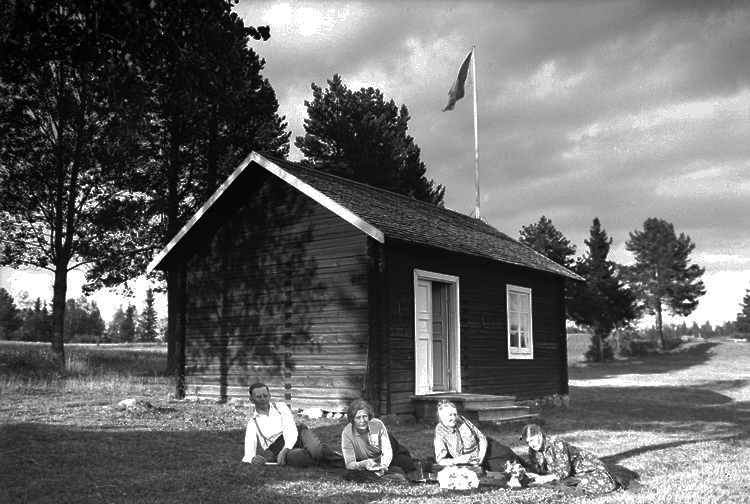 Några personer halvligger framför en stuga. Bakom stugan finns en ...