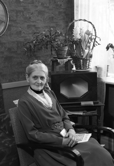 Foto av en äldre kvinna som sitter i en karmstol framför en radio. Fru ...