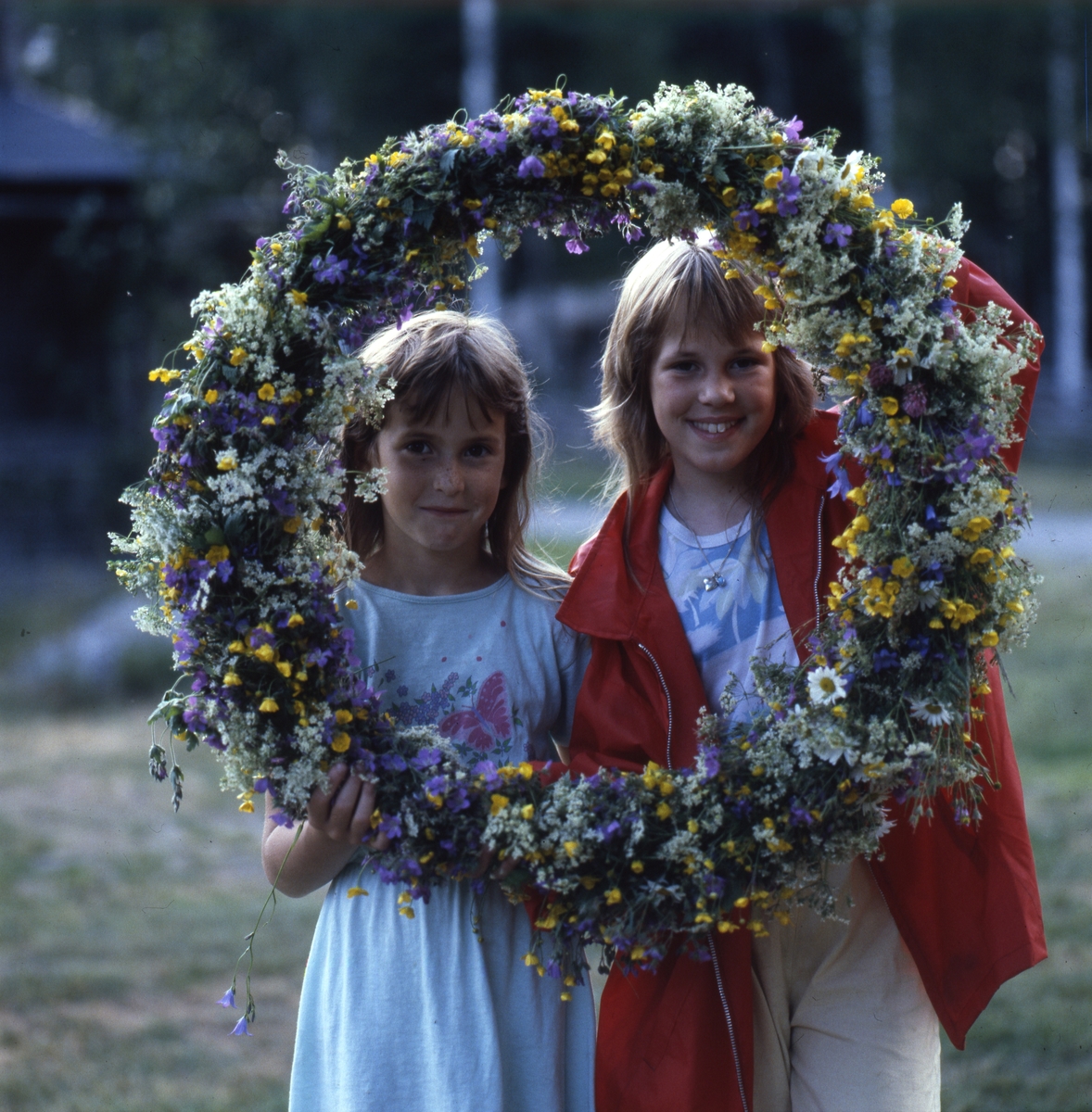 Två flickor tittar fram igenom en stor blomkrans. Troligen är den till ...