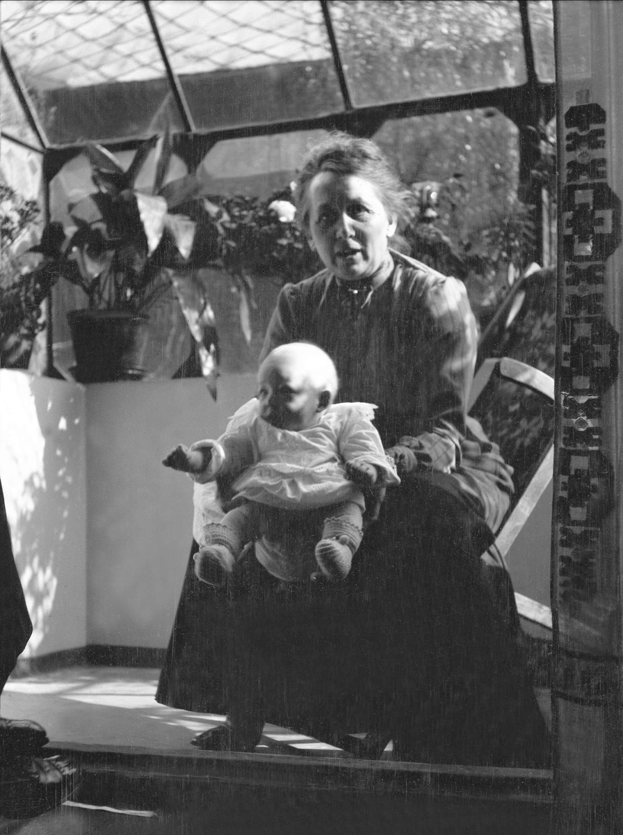 En kvinne sitter på en stol i en innglassert balkong med en baby på ...