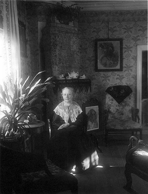 Porträtt av Valborg Franck i sitt hem. Hon sitter i en stol framför en kakelugn. Ett gnistskydd ...