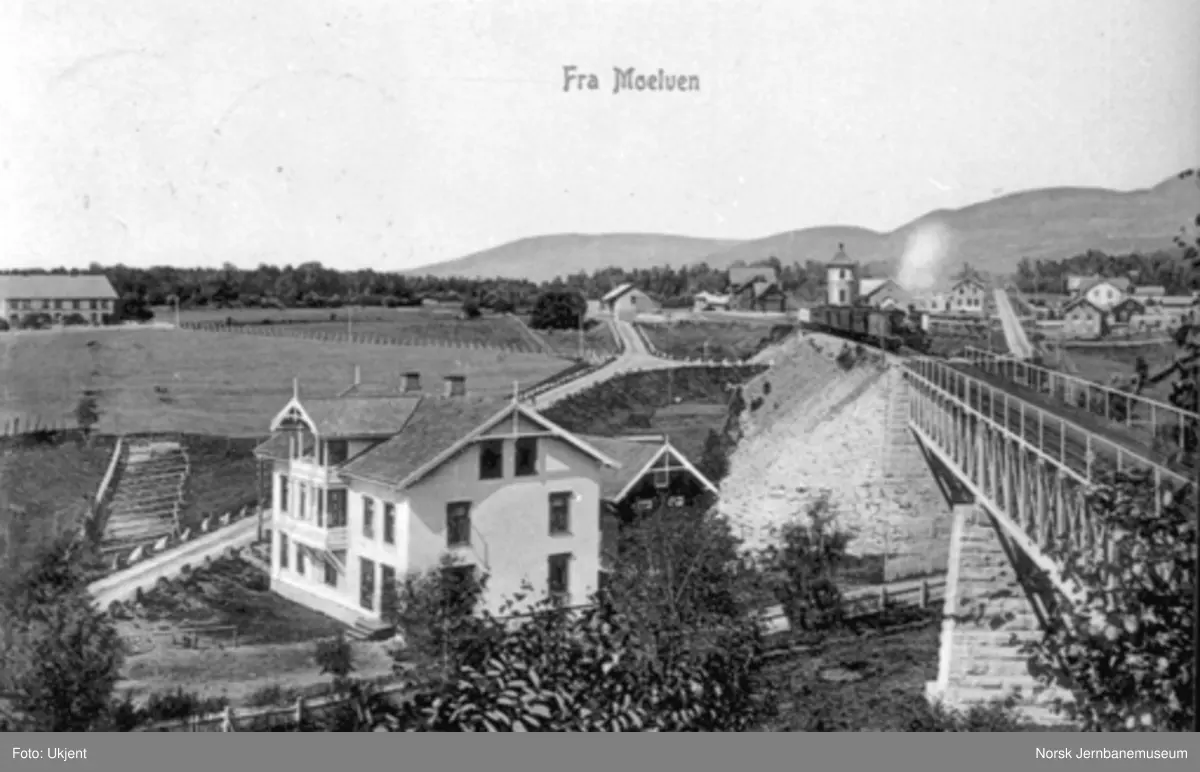 Moelv stasjon : oversiktsbilde med jernbanebrua og tog som forlater ...